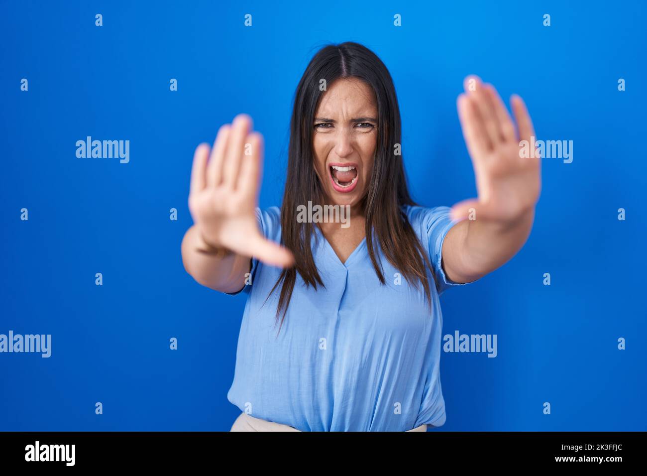 Young brunette woman standing over blue background doing stop gesture ...
