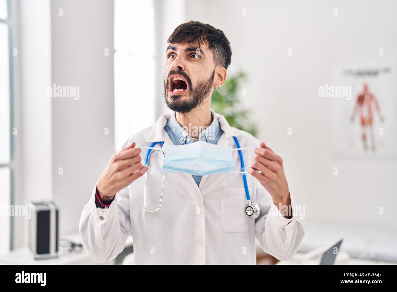 Young hispanic doctor man with beard holding safety mask angry and mad ...