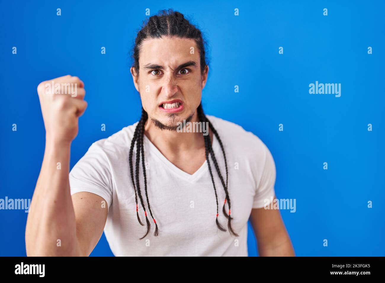 Hispanic man with long hair standing over blue background angry and mad ...