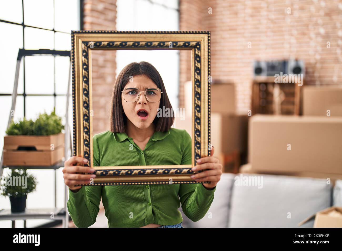 Young hispanic woman at new home holding empty frame in shock face ...
