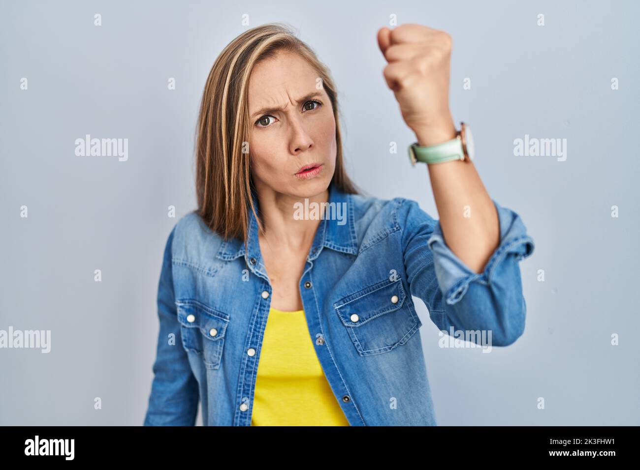 Young blonde woman standing over blue background angry and mad raising ...