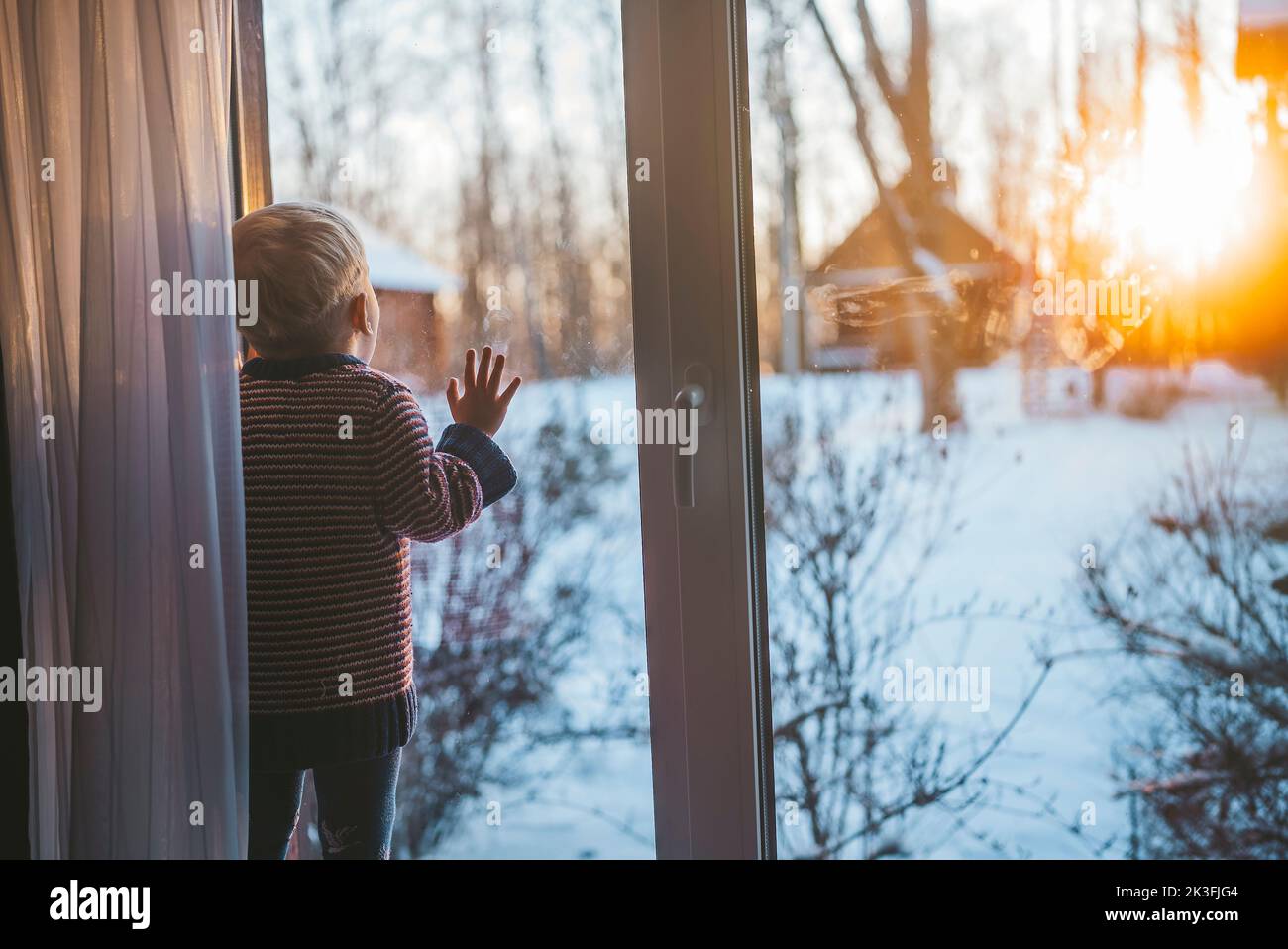 little boy looks out the window at home in winter waving his hand and ...