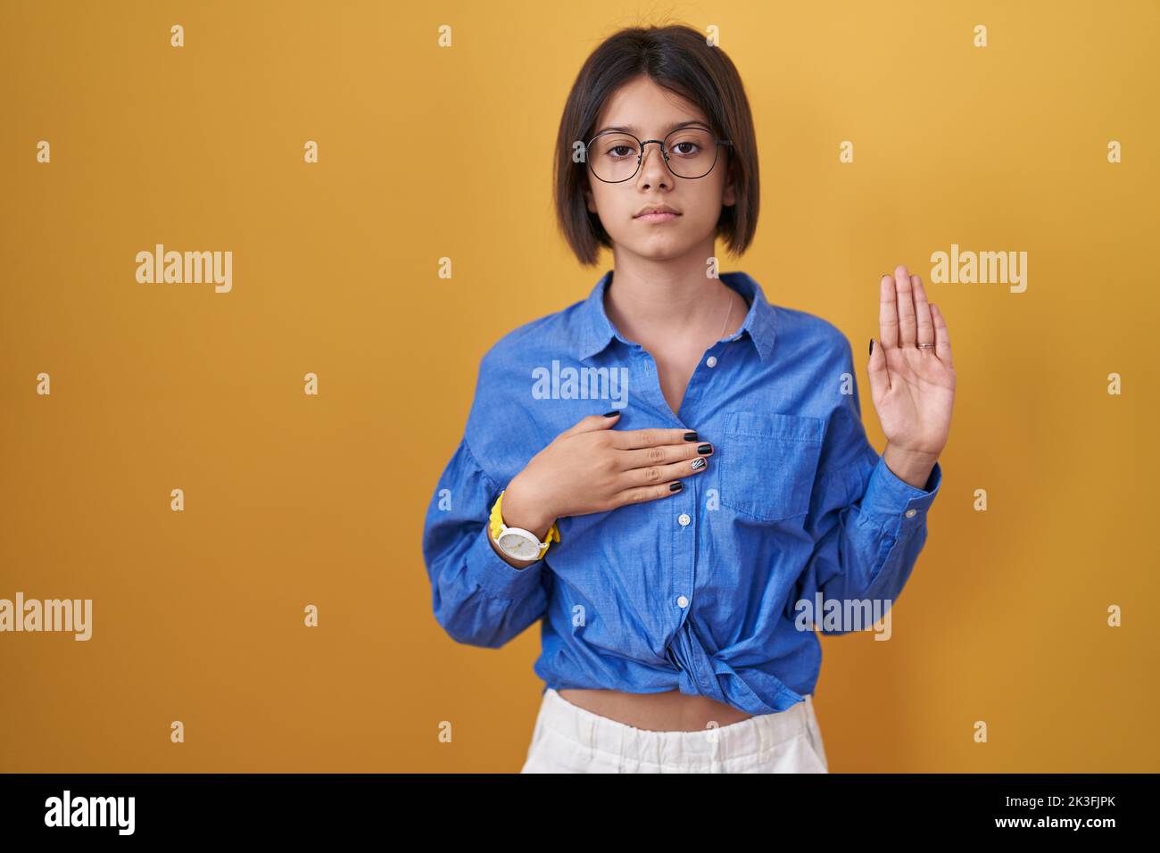 Young girl standing over yellow background swearing with hand on chest ...