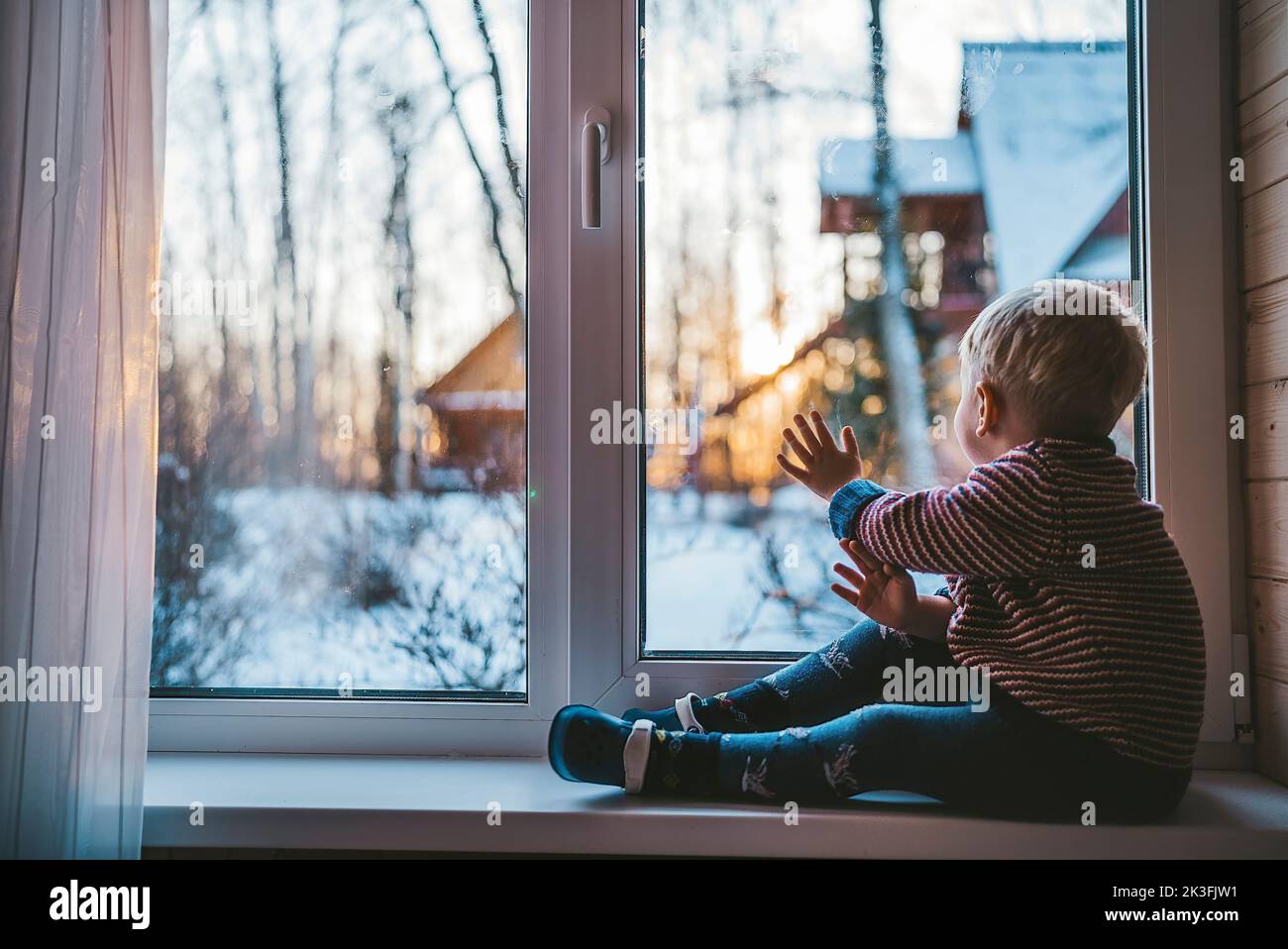 little boy looks out the window at home in winter waving his hand and ...