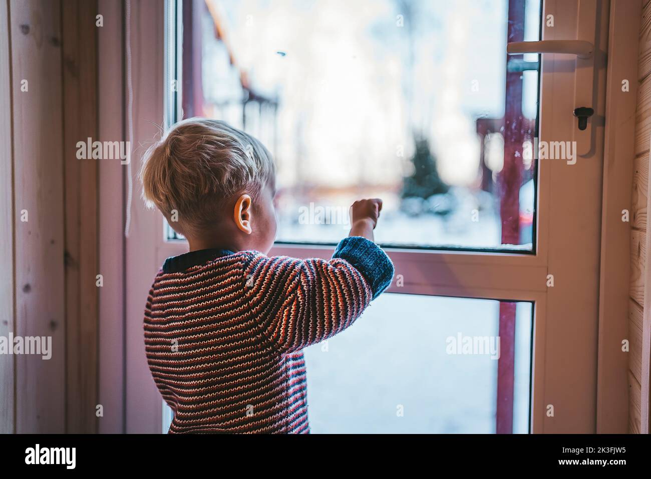 little boy looks out the window at home in winter waving his hand and ...