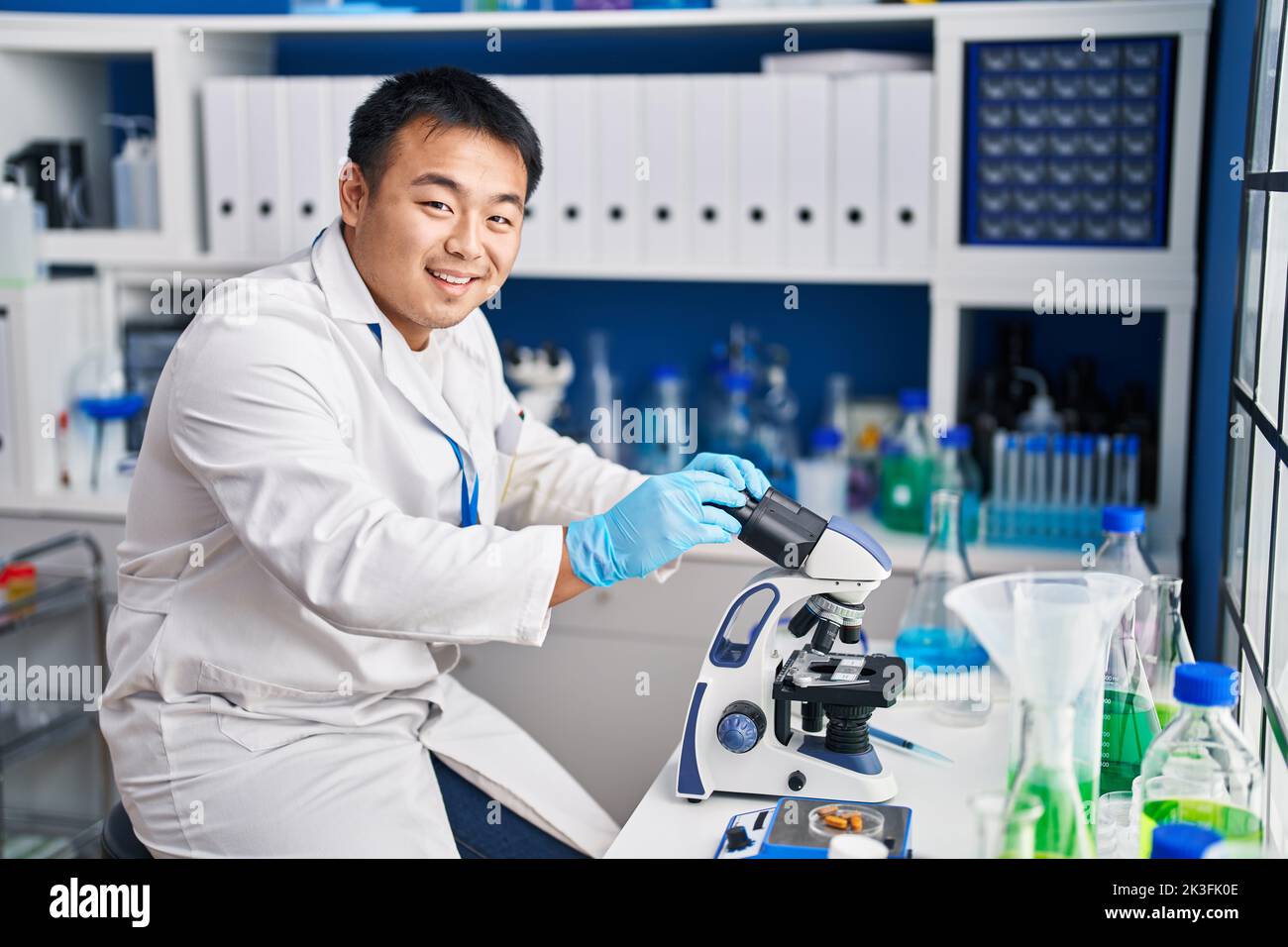 Young chinese man wearing scientist uniform using microscope at ...