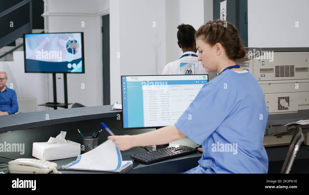Medical assistant working on appointments at registration desk in facility lobby. Woman nurse ...