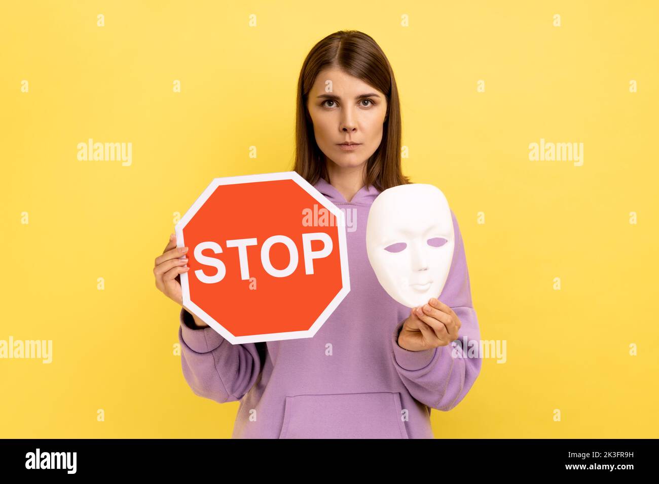 Portrait of serious woman holding red stop sign and white mask, looking ...