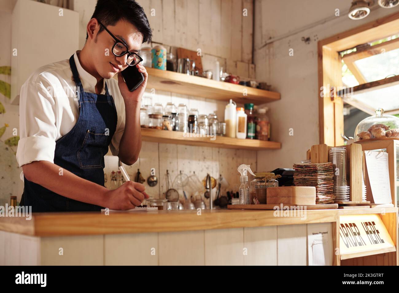 Coffeeshop waiter talking on phone with customer and writing down order ...