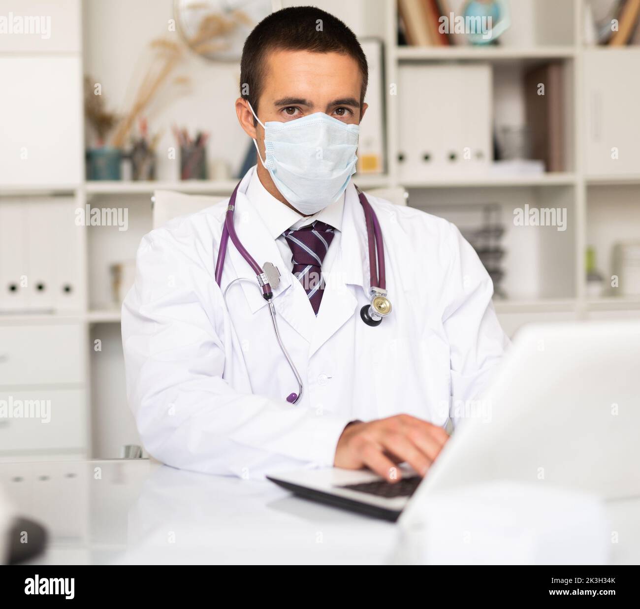 Doctor in protective medical mask sitting at workplace with computer in ...