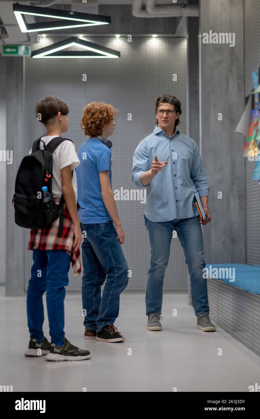 Serious schoolteacher conversing with pupils in the school corridor ...