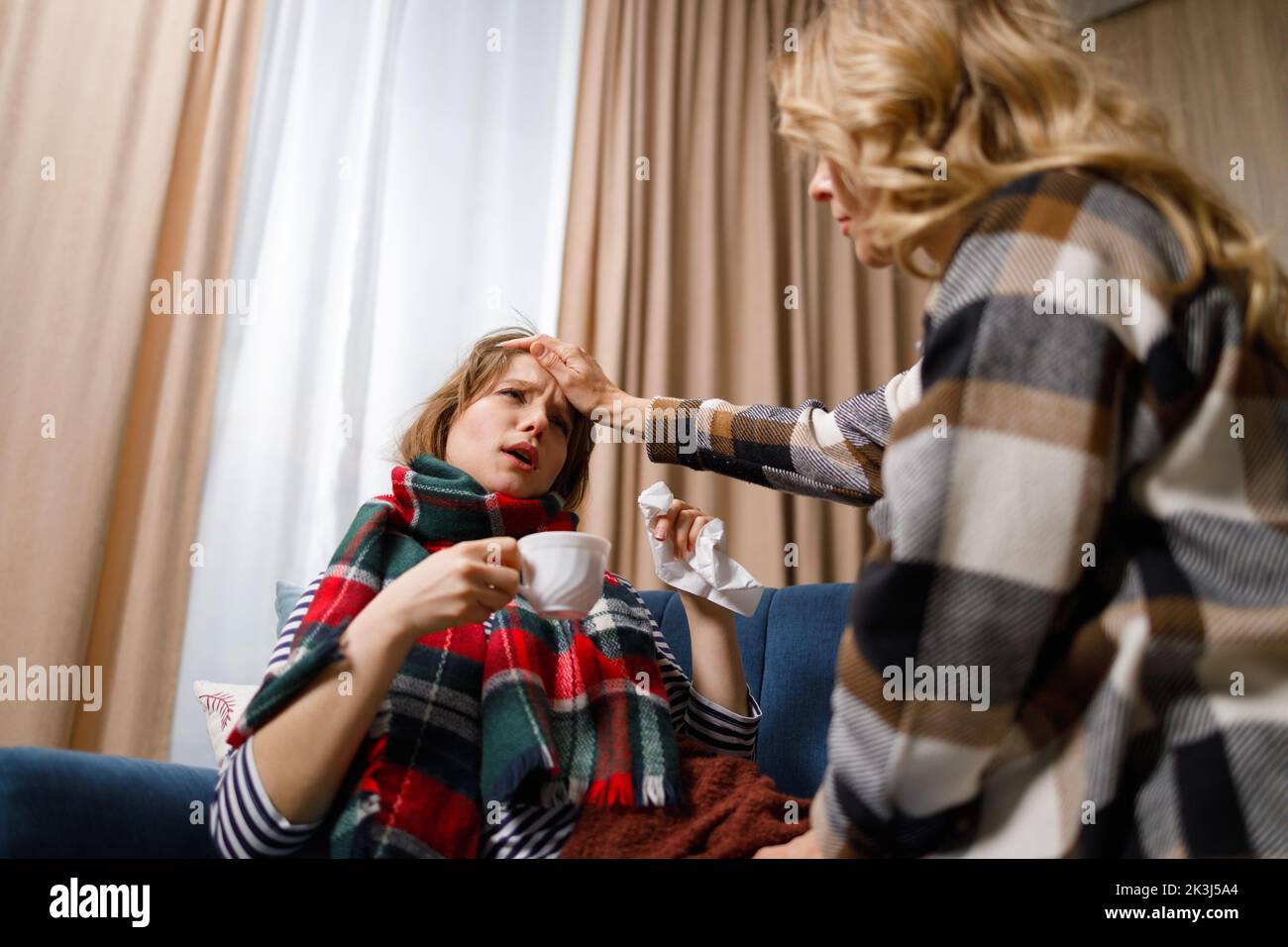 Girl with the flu drinks hot tea while her mother checks her forehead ...