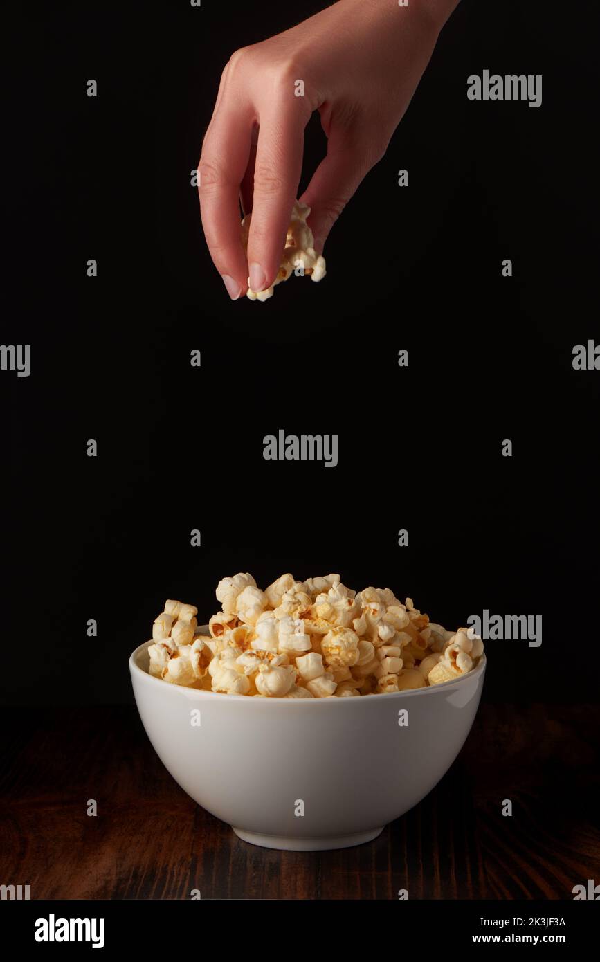 Woman’s hand taking popcorn from full white porcelain bowl on dark ...