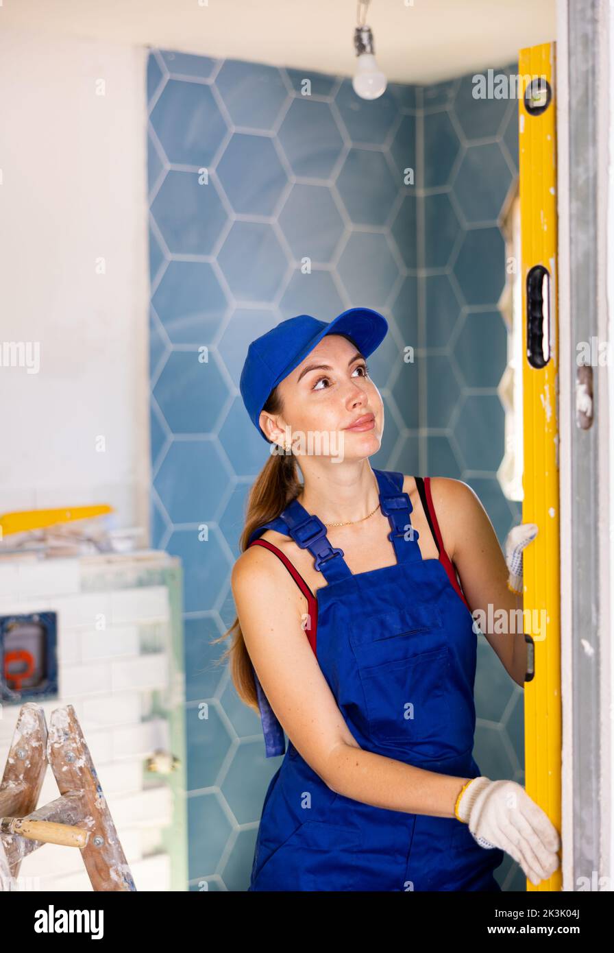 Pretty young woman tiler in blue construction overalls and cap using ...