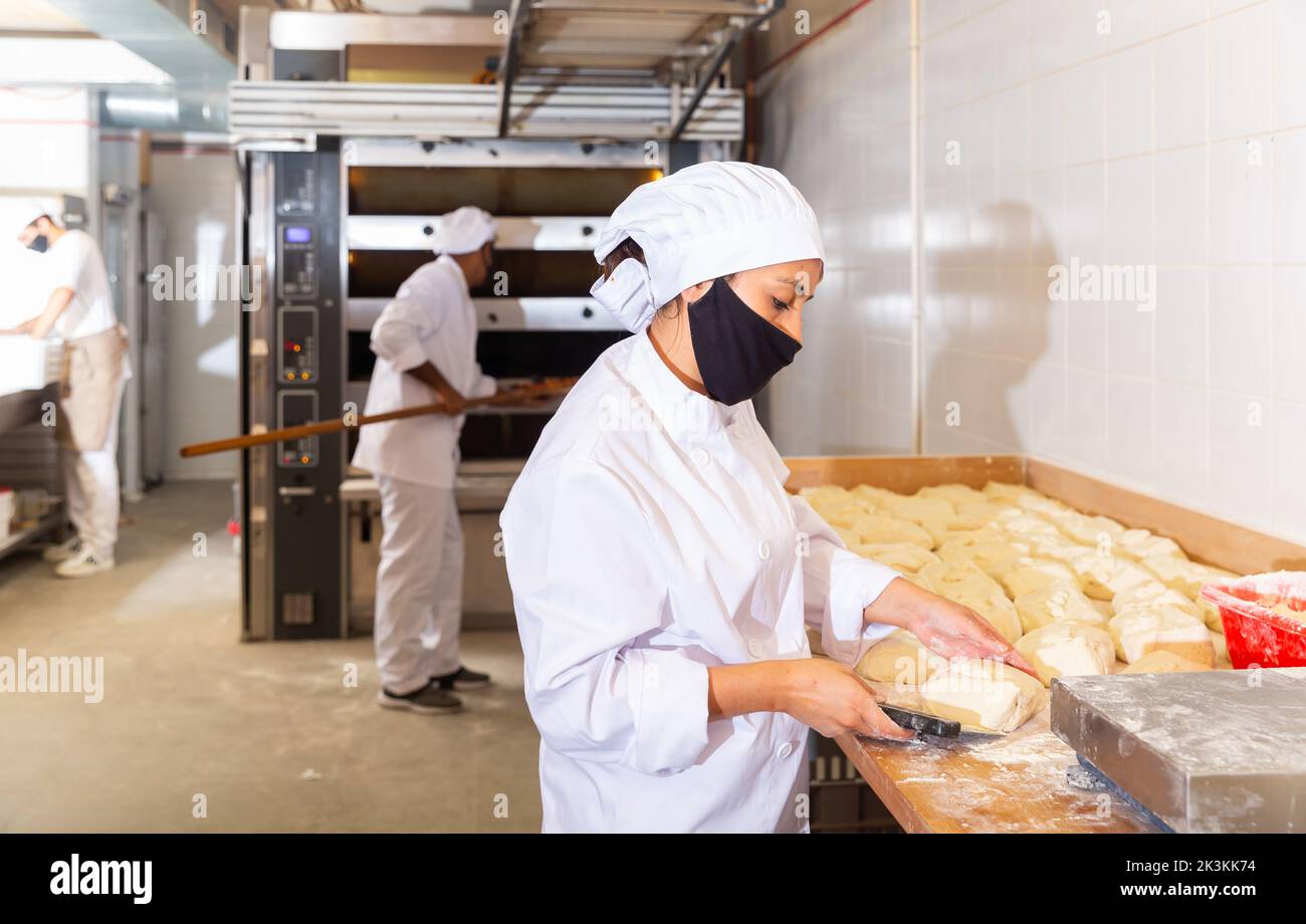 Female bakery worker wearing protective mask cuts dough with knife into ...