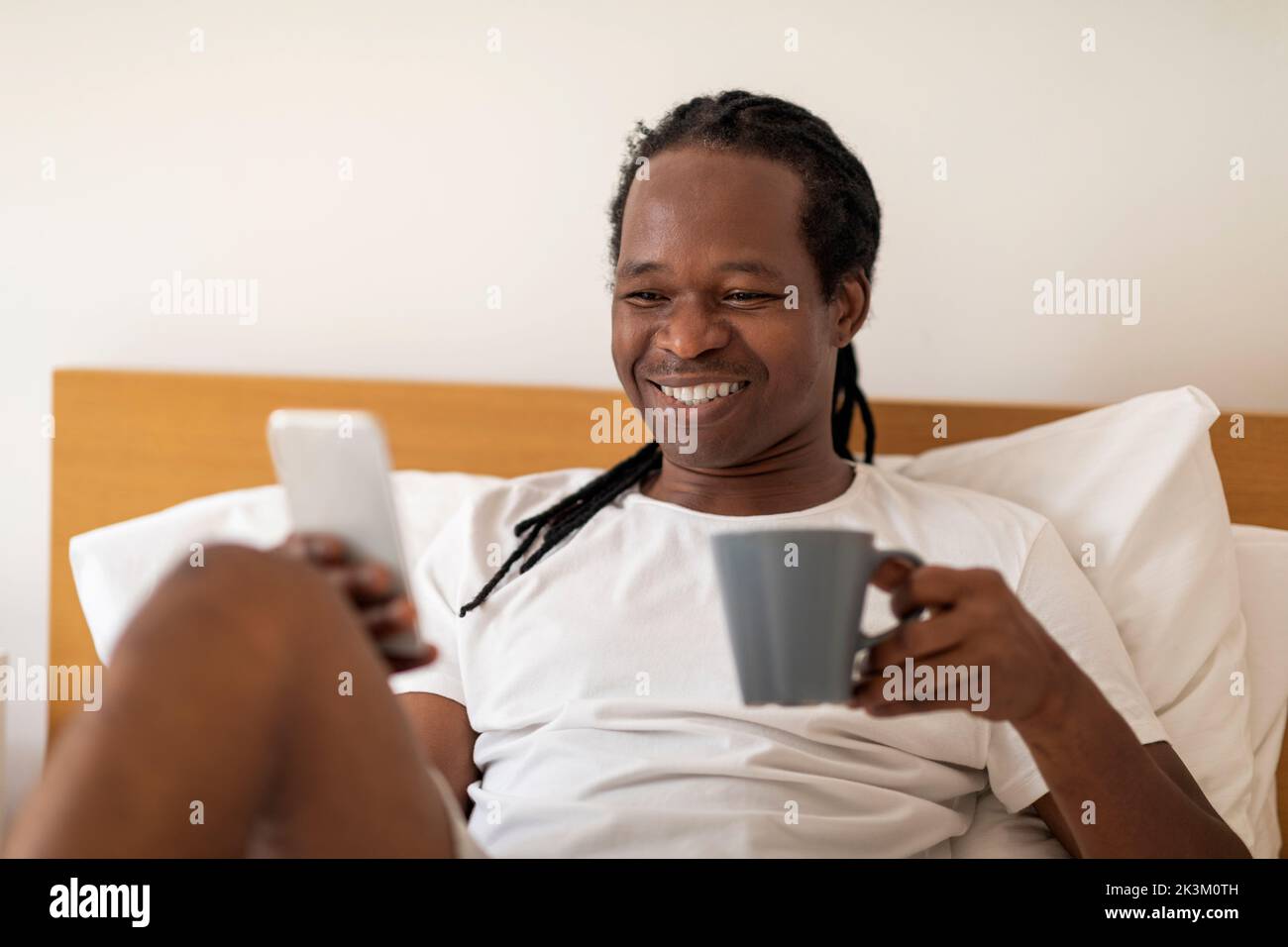 Smiling Young Black Man Resting In Bed With Smartphone And Coffee Stock ...