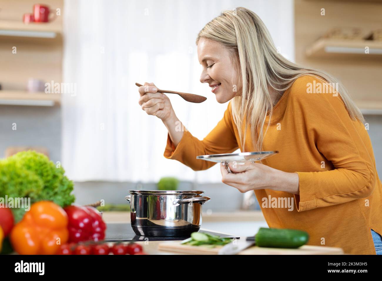Pretty middle aged woman tasting food while cooking, kitchen interior ...