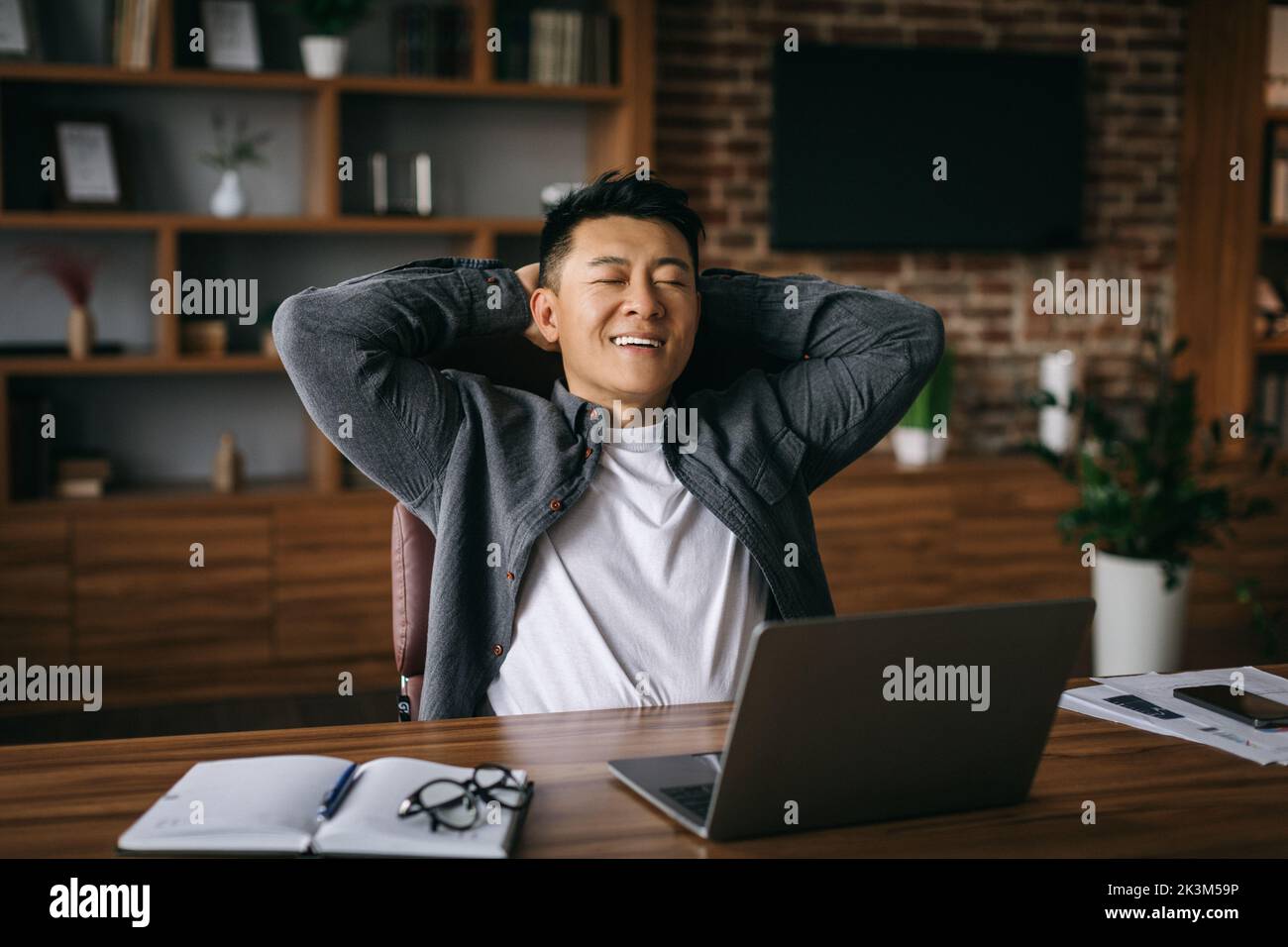 Smiling adult chinese male enjoys rest in chair at table with computer ...