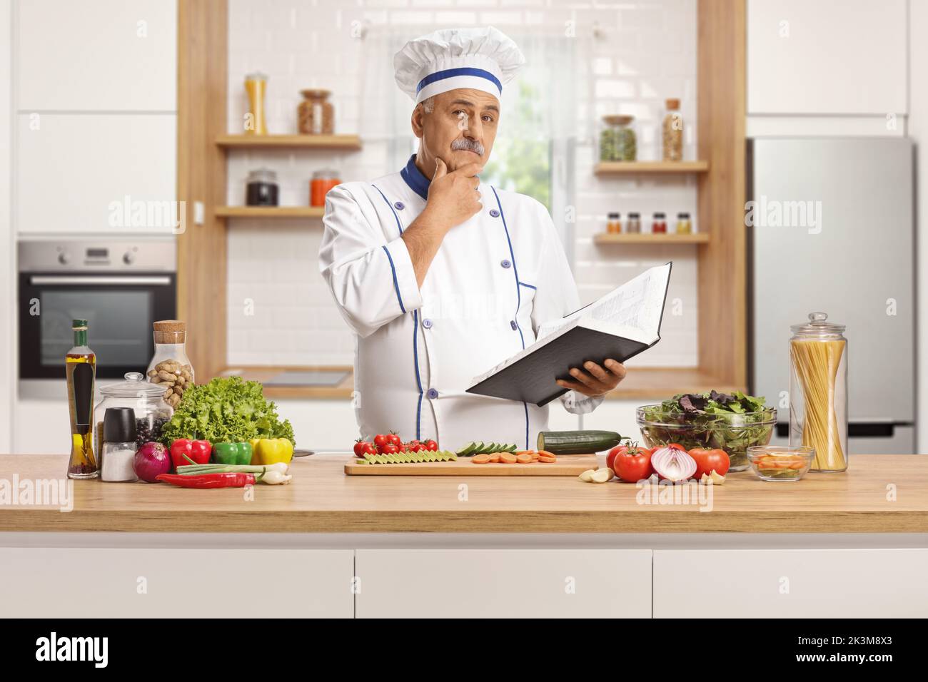 Mature male chef standing behind a kitchen counter holding a cook book ...