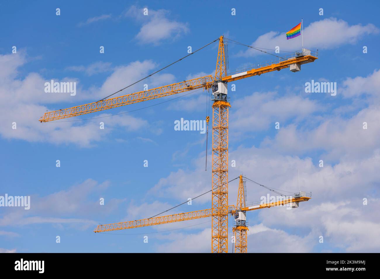 A pair of yellow cranes with a big Pride flag against the cloudy blue ...