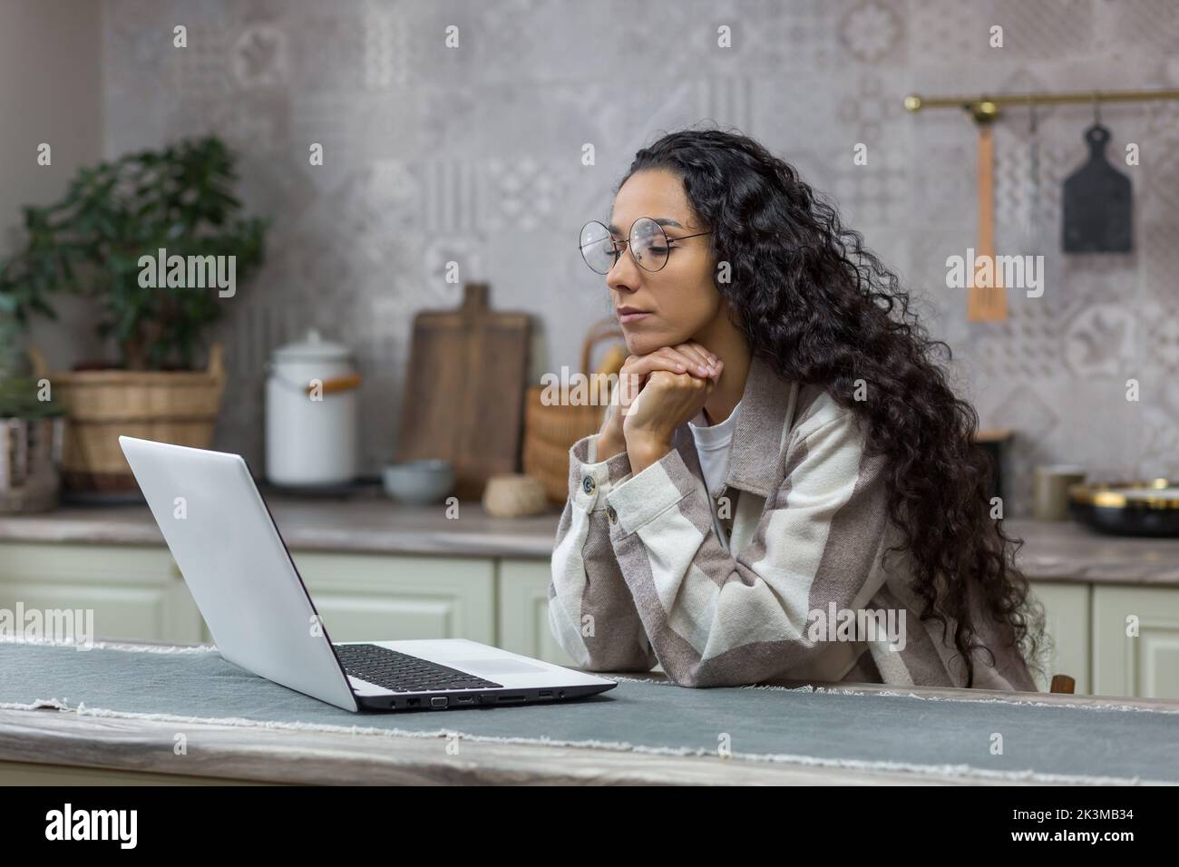 Hispanic woman thinking at home sitting in kitchen working on laptop ...
