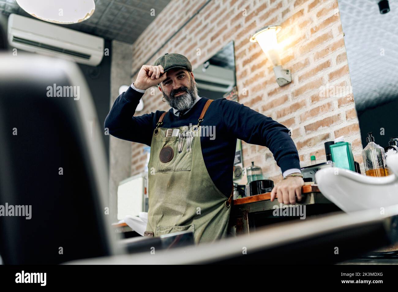Low angle of cheerful mature male barber wearing cap and apron standing ...