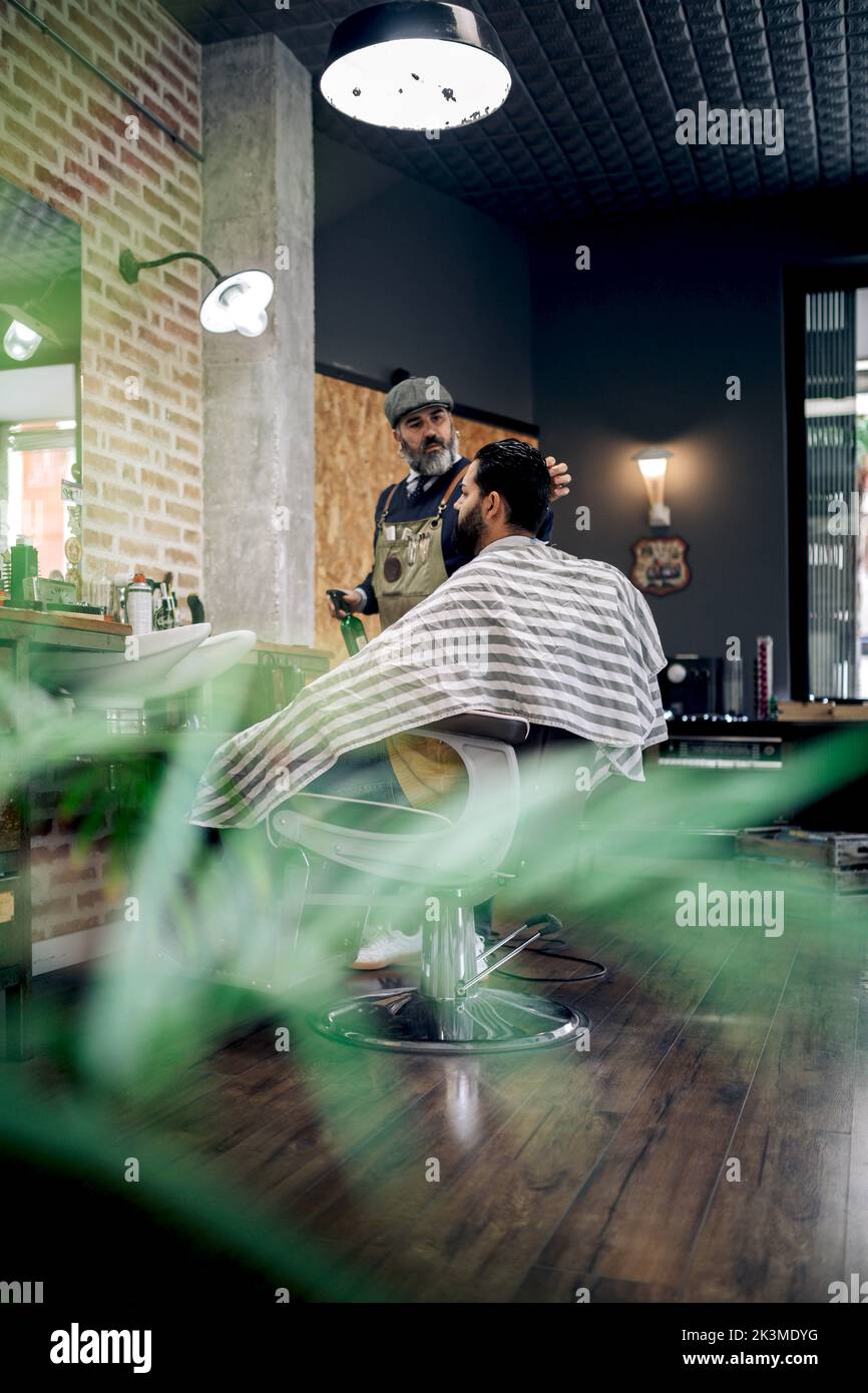 Side view full body of bearded barber spraying hair of male customer ...