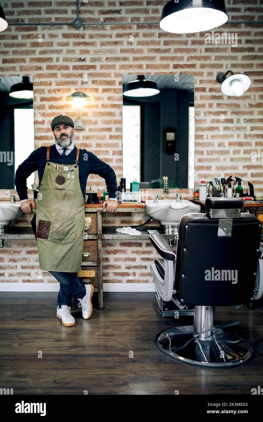 Full body of middle aged male barber in cap and apron standing near ...