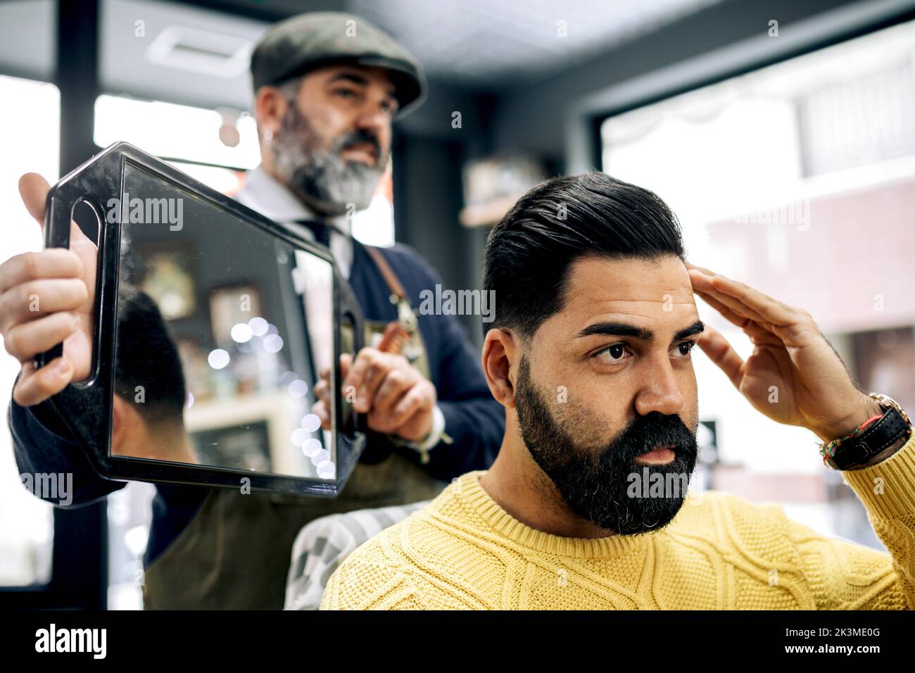 Positive mature bearded ethnic male barber with mirror in hand smiling ...