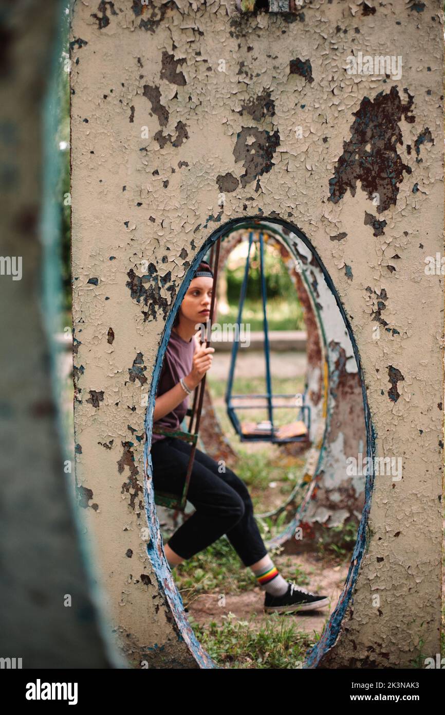 Young punk woman sitting on a swing at abandoned playground in summer ...