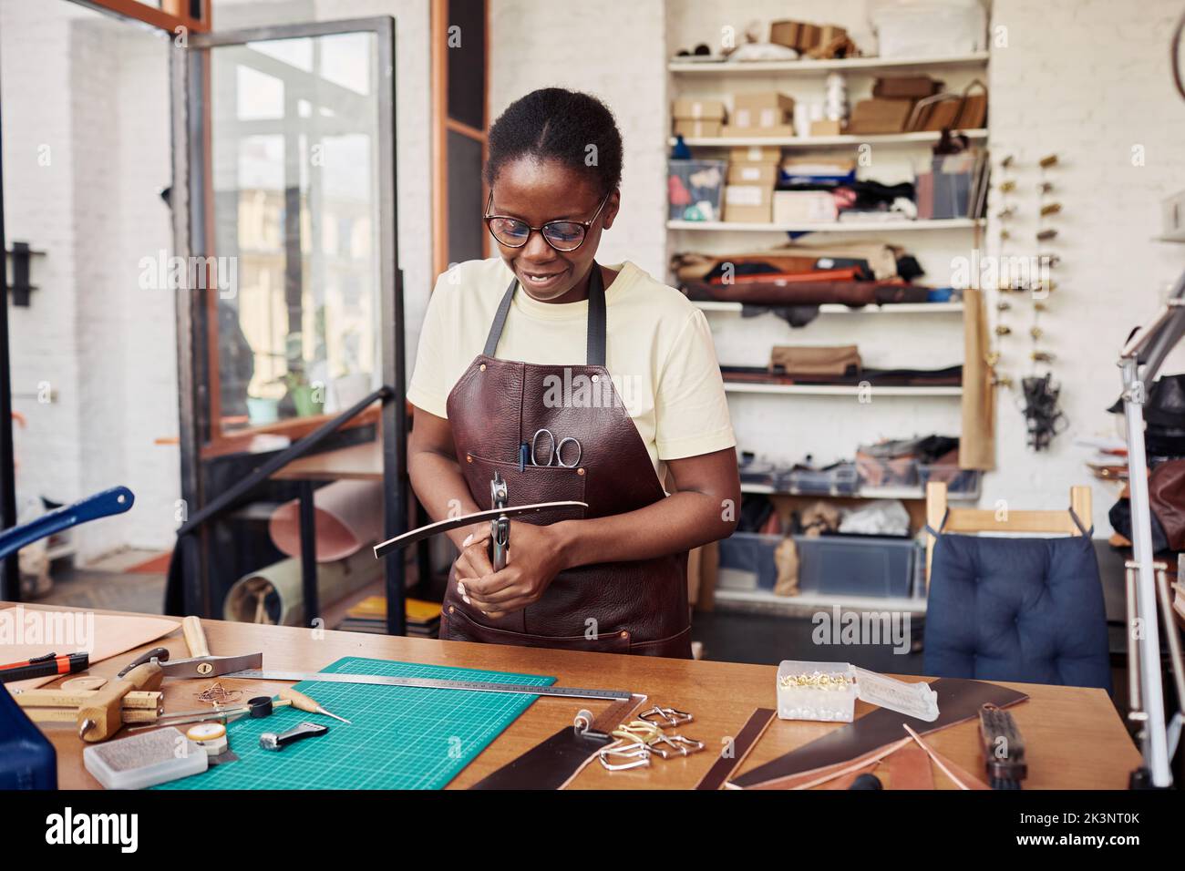 Waist up portrait of black female artisan punching holes in handmade belt in leatherworking shop ...