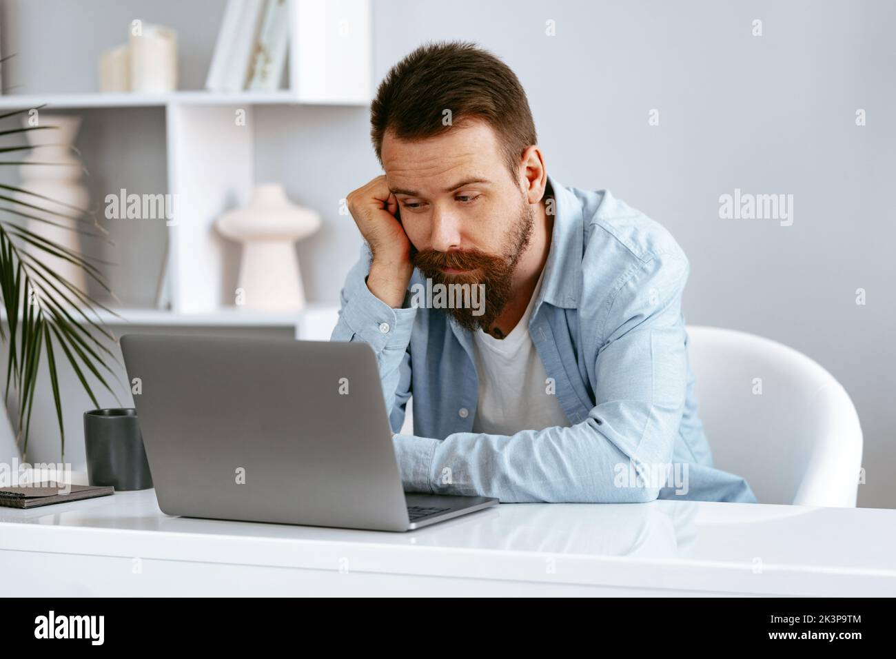 Thoughtful tired young bearded man sitting at the table working on ...