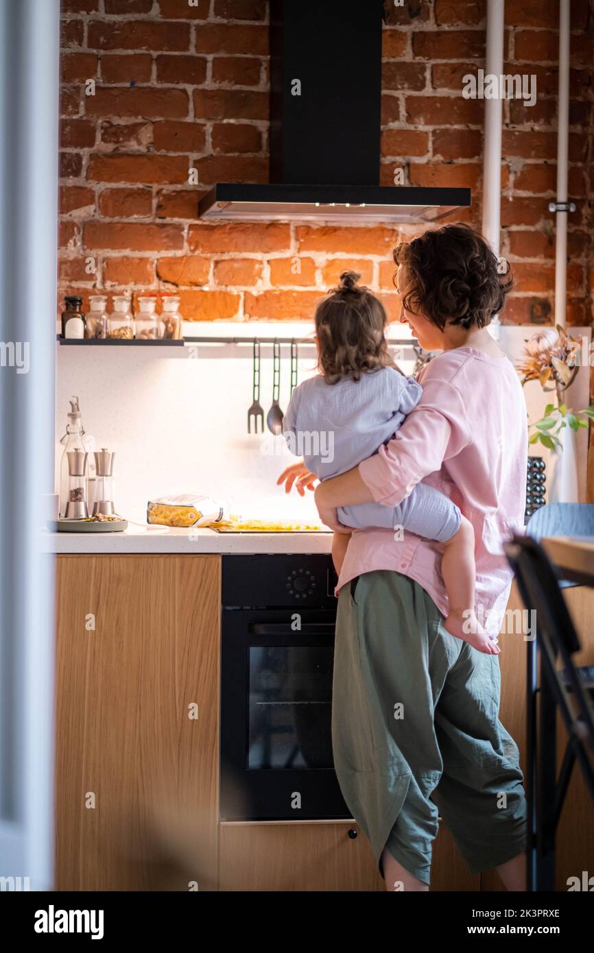 Young mother with baby girl on her hands cooking together at kitchen at ...