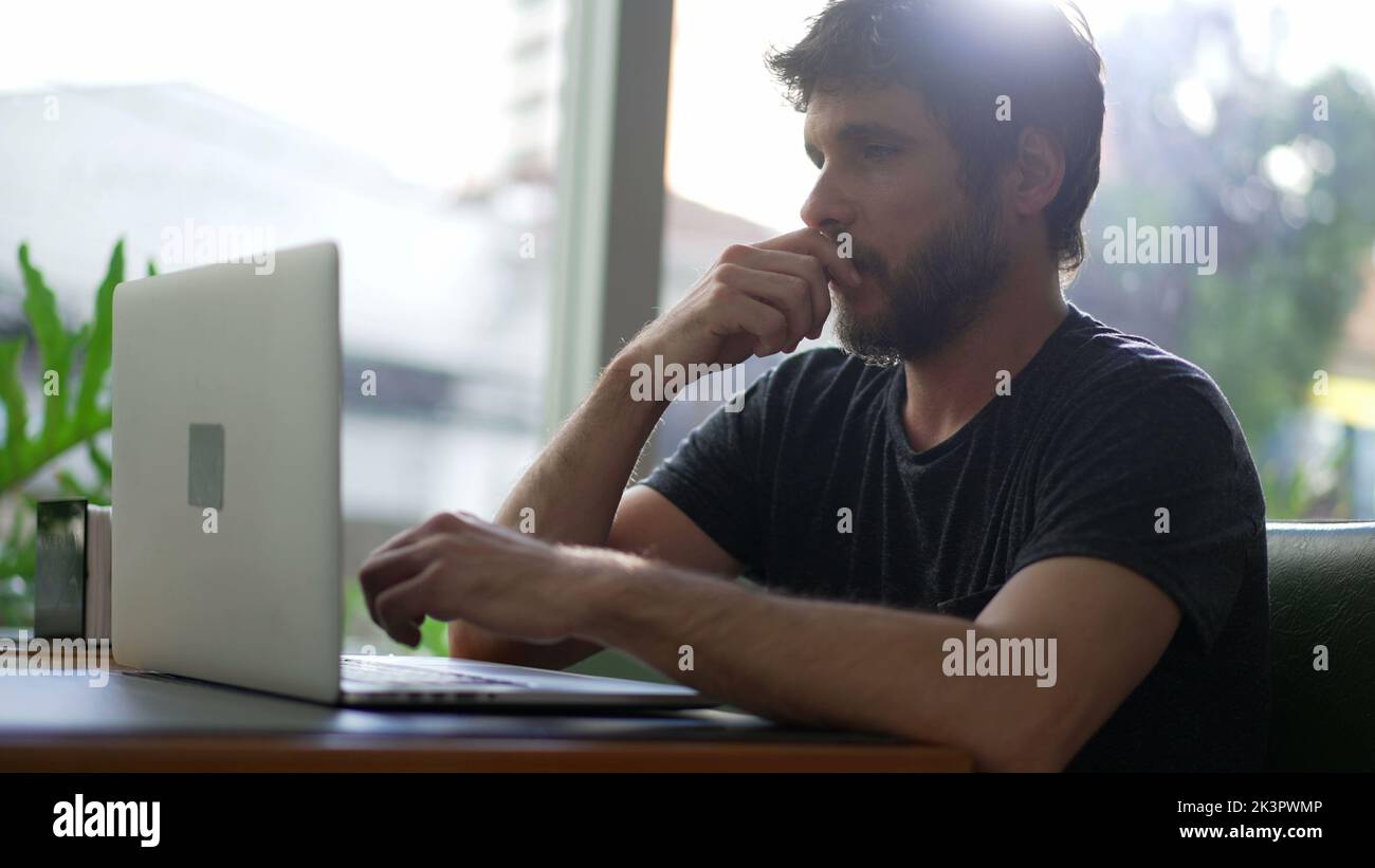 Thoughtful young man seated at coffee shop in front of laptop looking ...