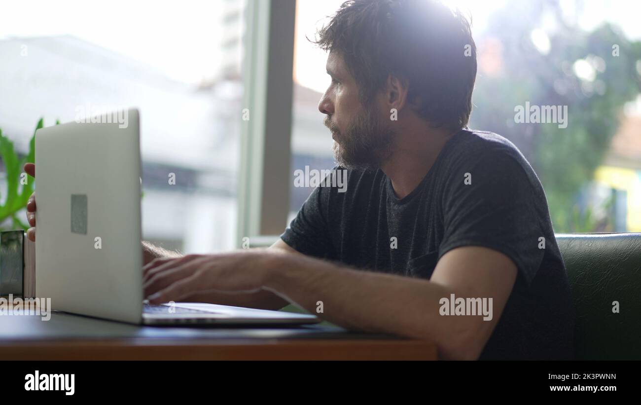 Thoughtful young man seated at coffee shop in front of laptop looking ...