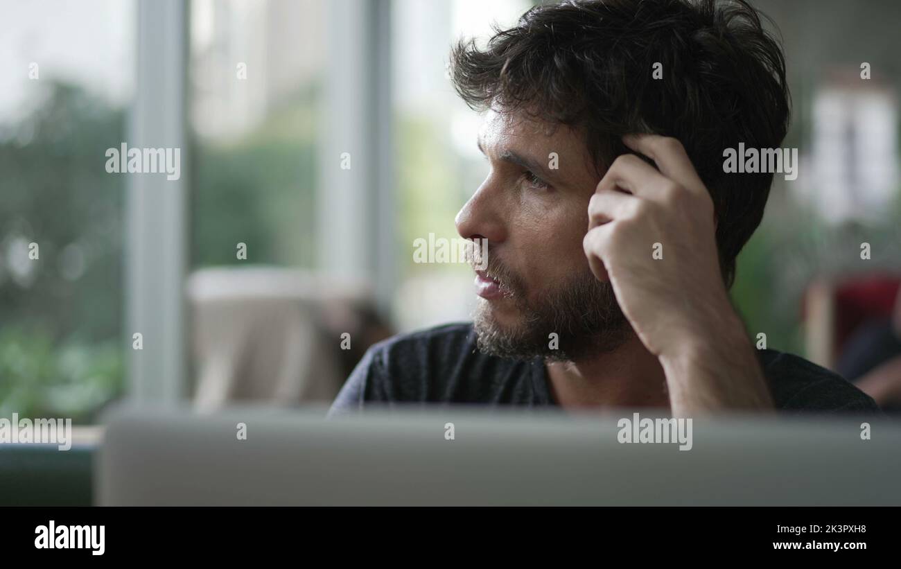 Pensive young man staring out the window in front of laptop computer ...