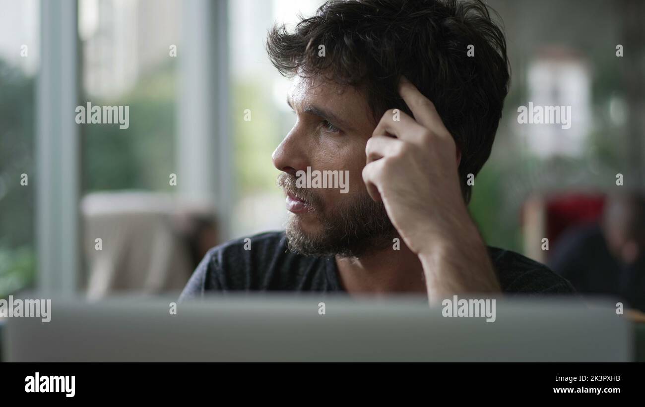 Pensive young man staring out the window in front of laptop computer ...