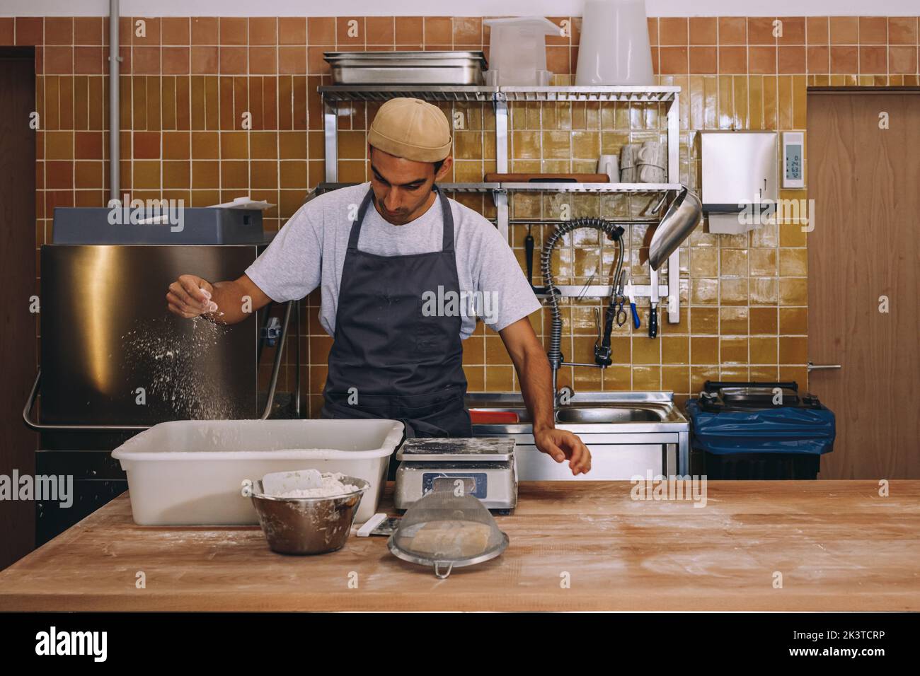 Positive young ethnic male baker in apron and hat sprinkling four on ...