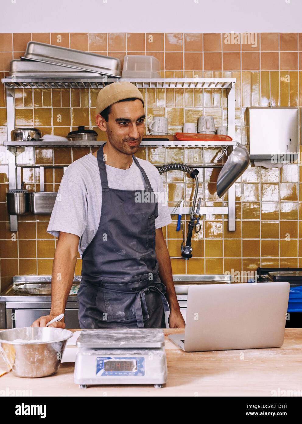 Positive young ethnic male bakery owner standing at counter with laptop ...