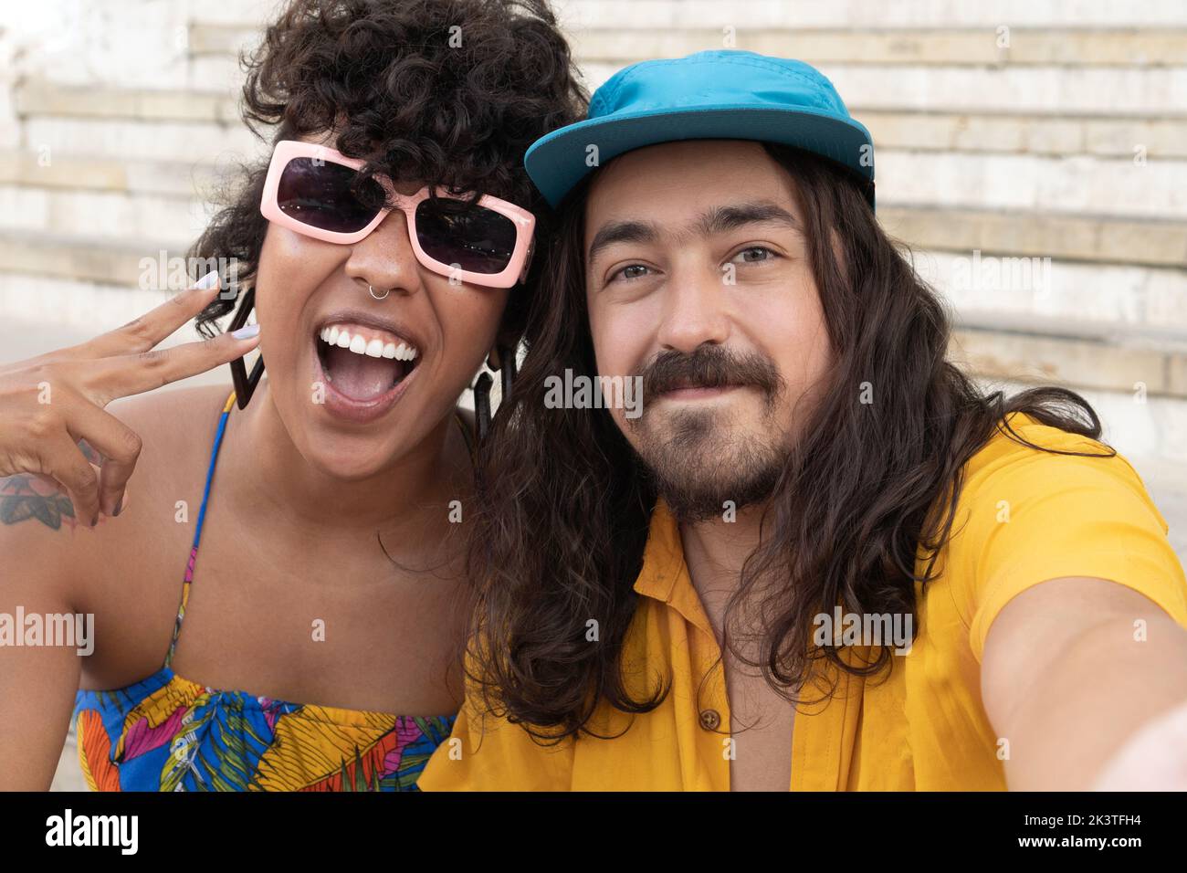 Positive ethnic couple of hipsters in colorful clothes showing v sign ...