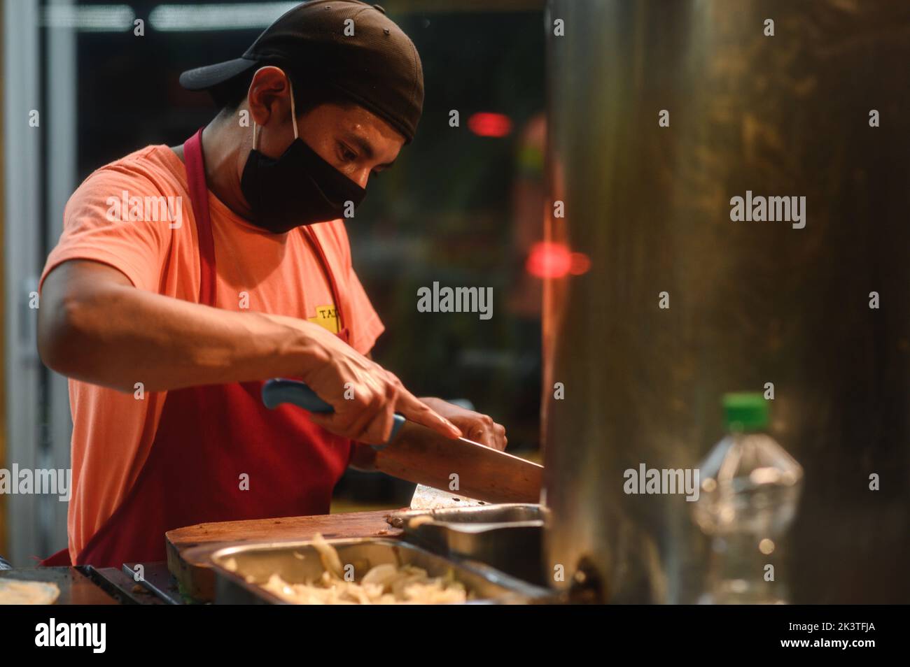 Ethnic male cook in fabric mask with cleaver cutting product at table ...