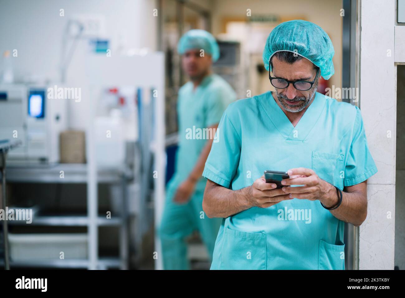 Man in surgeon uniform browsing modern smartphone while leaning on wall ...