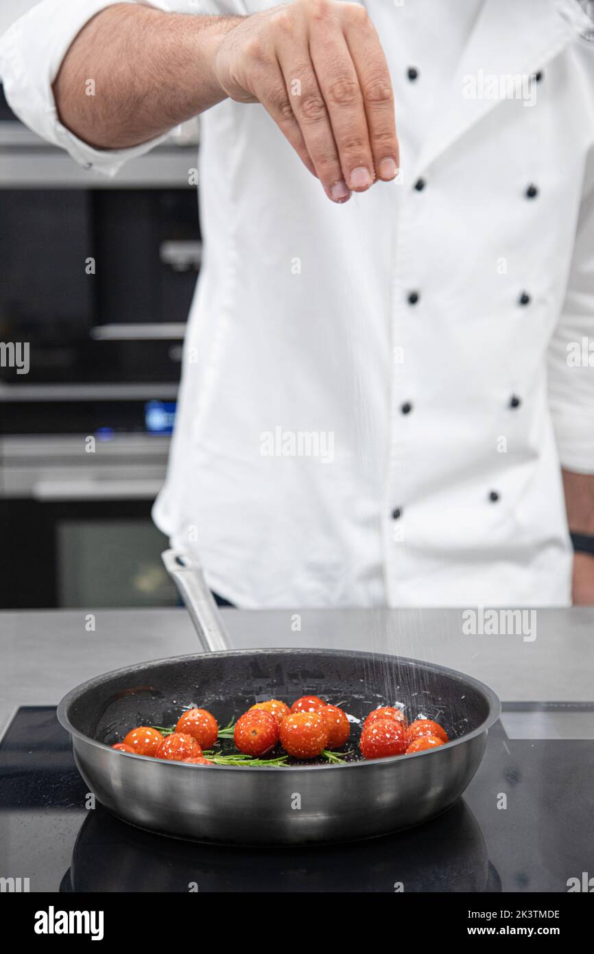 Crop man in white chef jacket adding pinch of salt to frying pan with ...