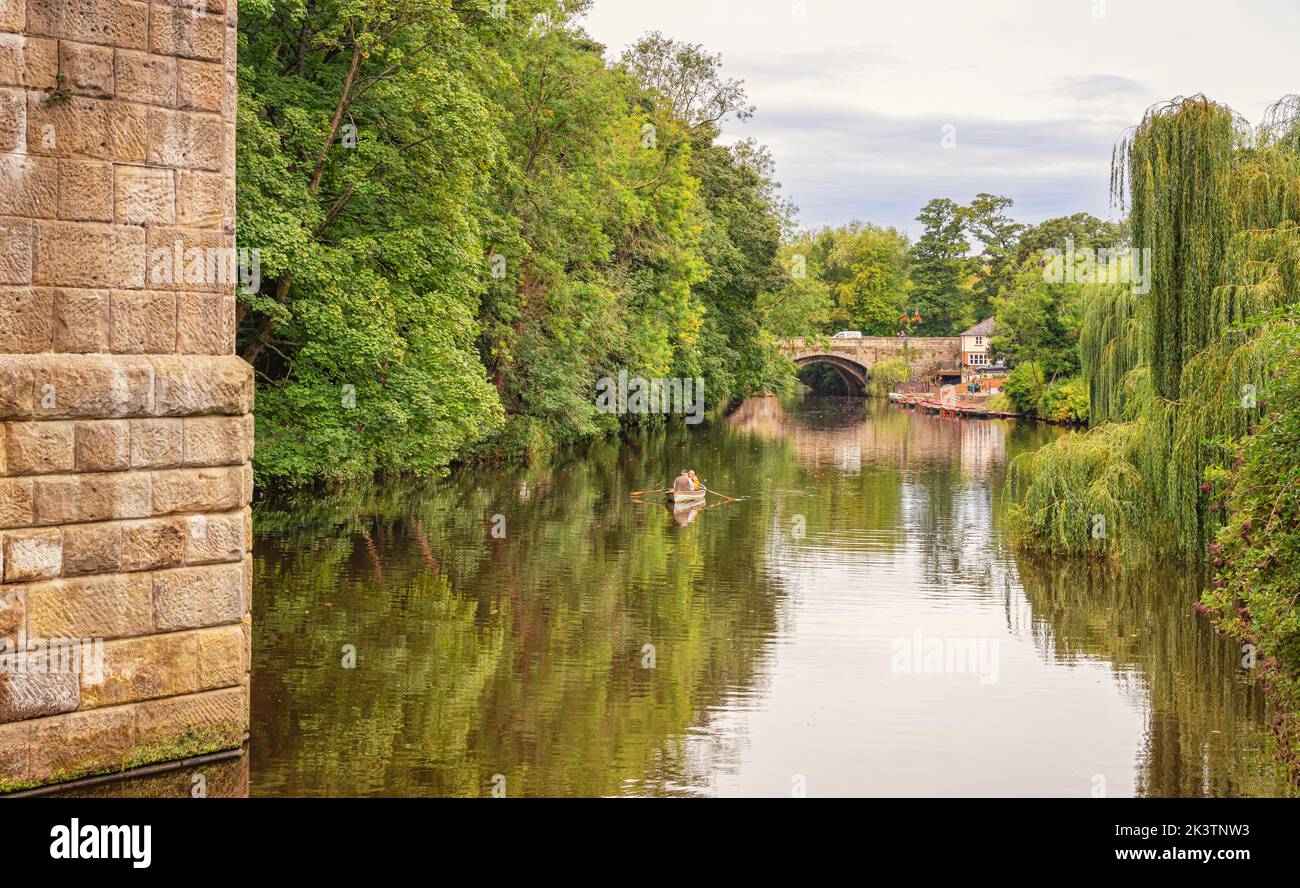A rowing boat on a river. A bridge is in the distance and trees line ...