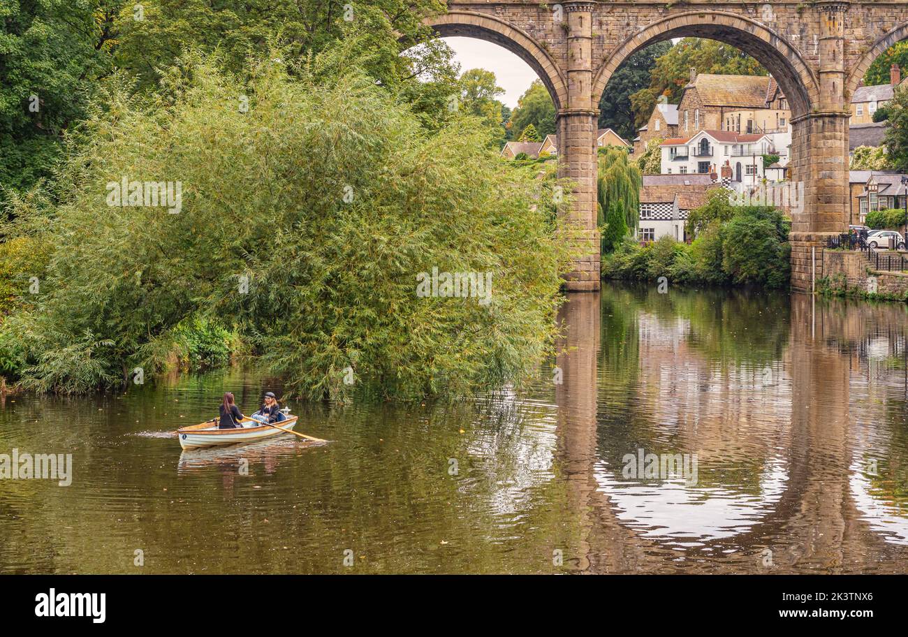 A rowing boat on a river with a historic viaduct behind. Trees are on ...