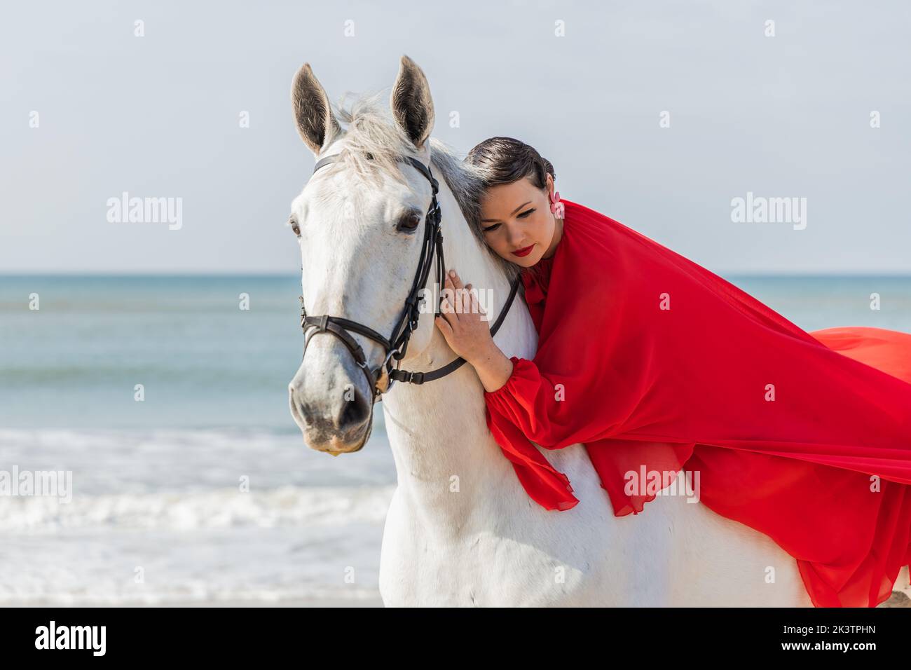 Side view of stylish female in red dress riding lying on purebred horse ...