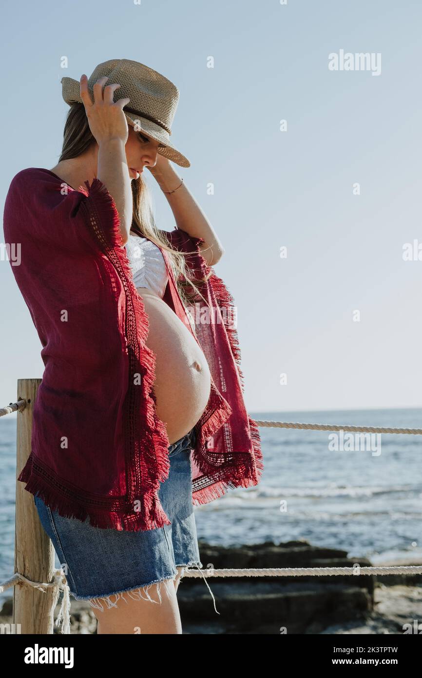 Pregnant female adjusting hat while standing near fence against sea and ...