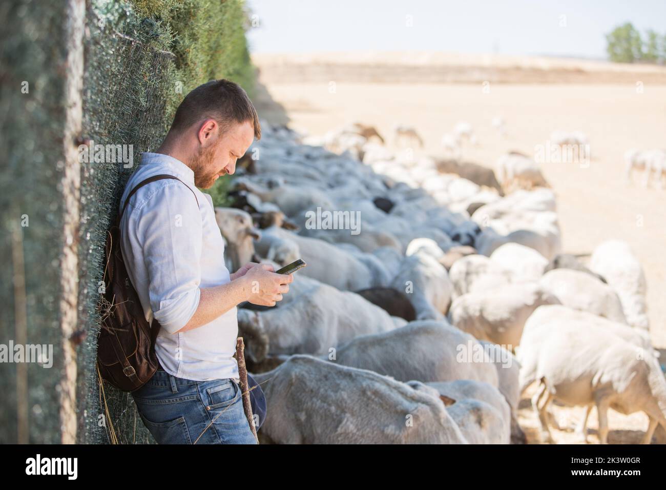 Side view of young shepherd with stick leaning on a wall resting while ...