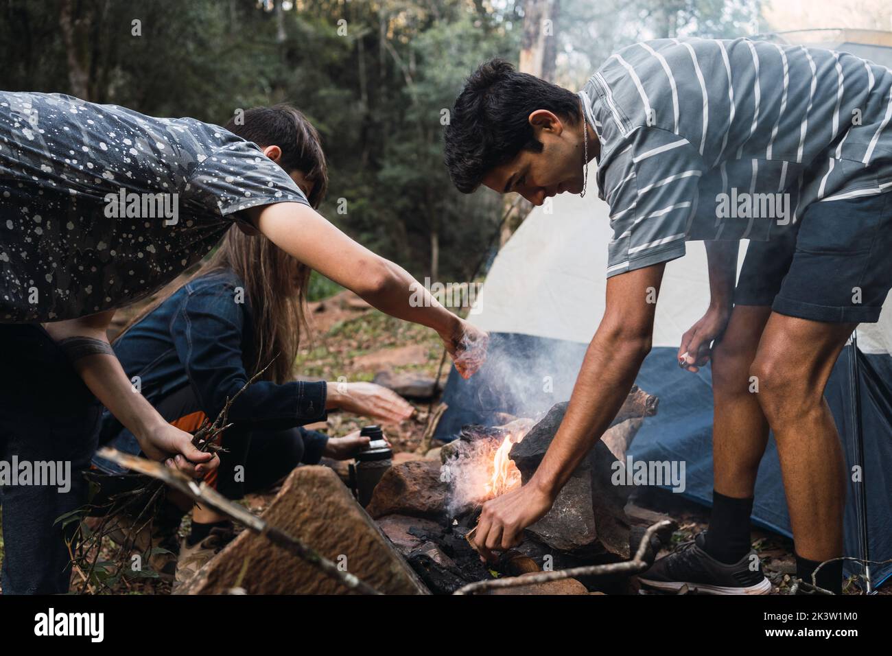 Side view of male hikers kindling fire with twigs near tent and bicycle ...
