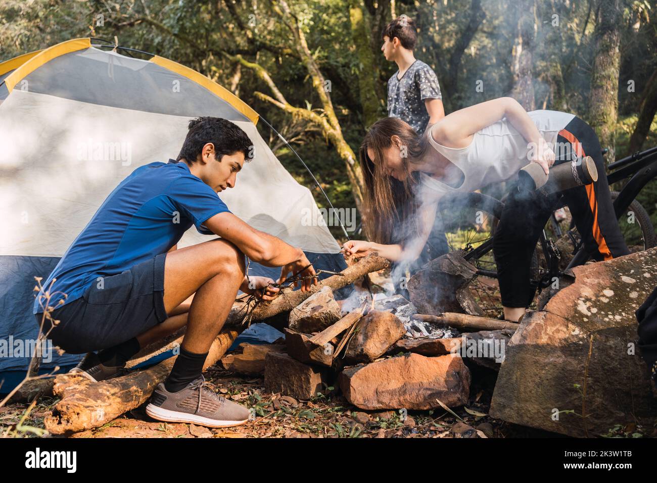 Multiethnic young explorers kindling smoking bonfire near tent during ...